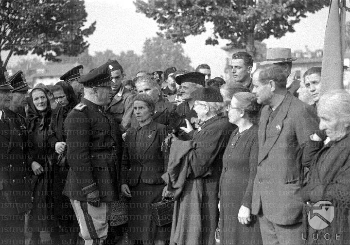 Benito Mussolini passa davanti a un gruppo di donne e uomini, in abiti scuri, famigliari di ...
